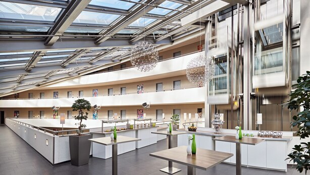 Um átrio espaçoso e moderno, com pé-direito alto e claraboias, apresentando vários níveis, mobiliário contemporâneo e elementos decorativos.