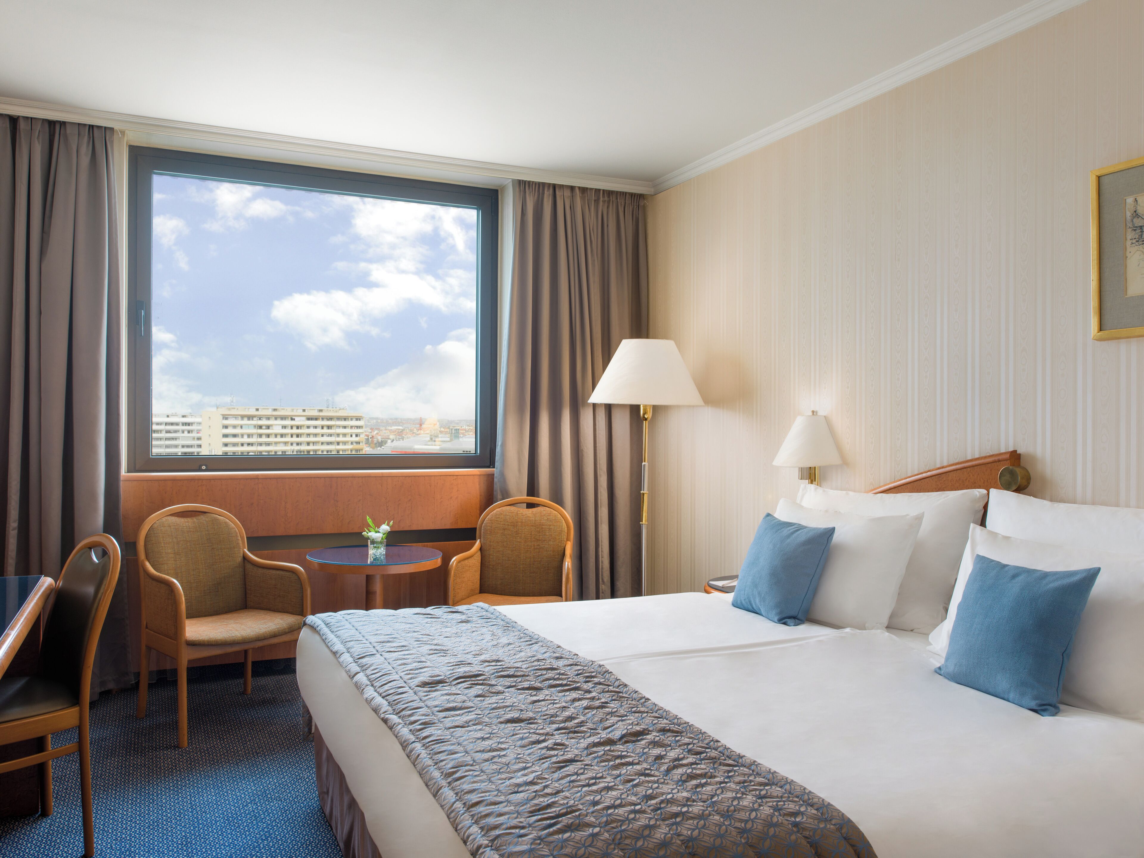 Quarto de hotel com uma cama grande, duas cadeiras, uma escrivaninha, um abajur e vista da cidade através de uma janela ampla.