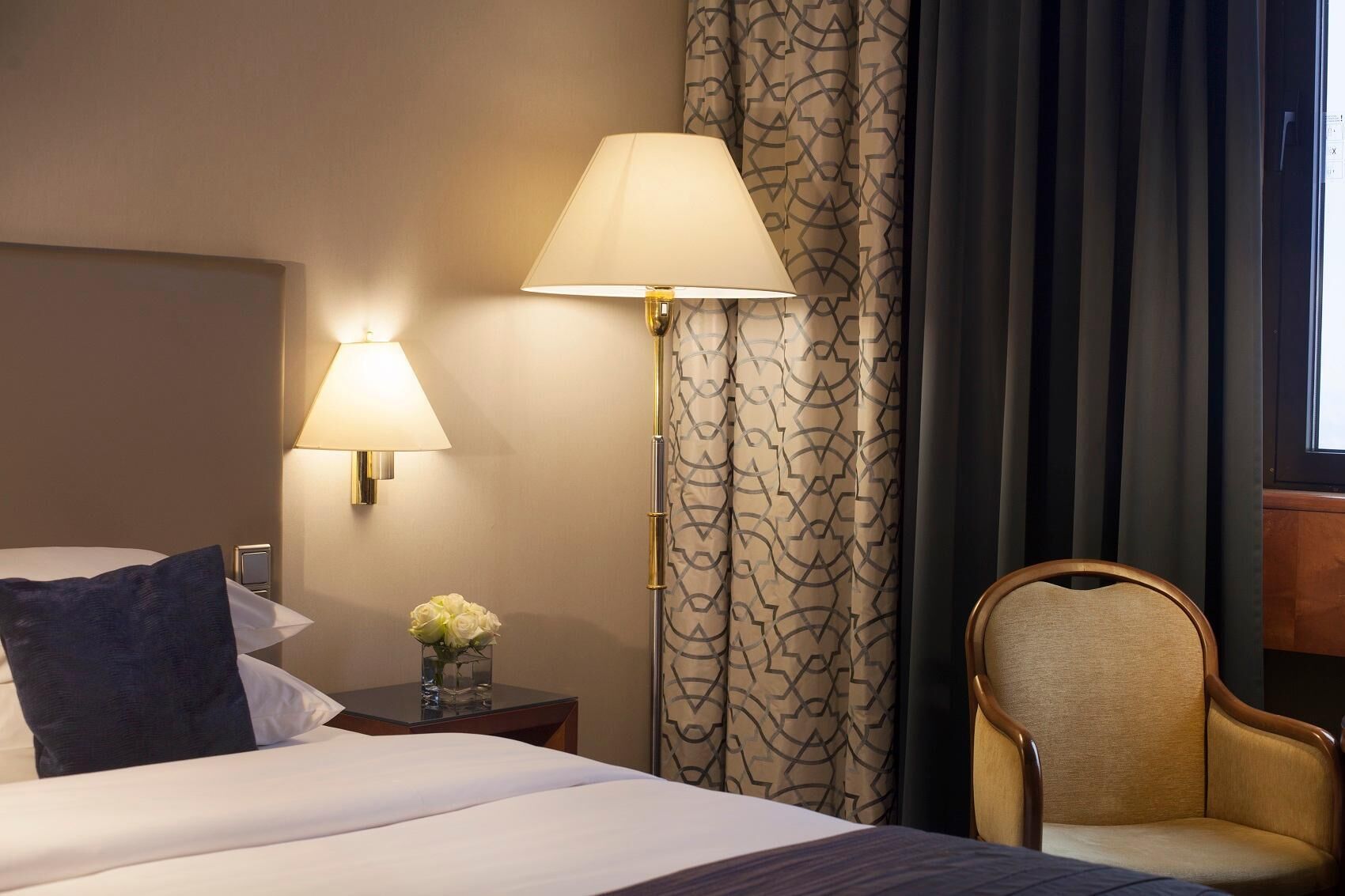 Quarto de hotel com uma cama, duas luminárias de parede, uma mesa de cabeceira com um vaso de flores, cortina estampada e uma cadeira de madeira.