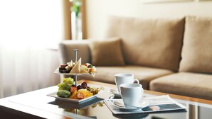 Uma mesa com um suporte escalonado para sobremesas, frutas e duas xícaras brancas, em um quarto com um sofá bege.