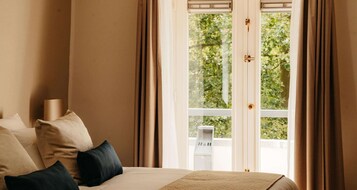 Quarto com cama, travesseiros, luminária e vista para o verde através de uma porta.