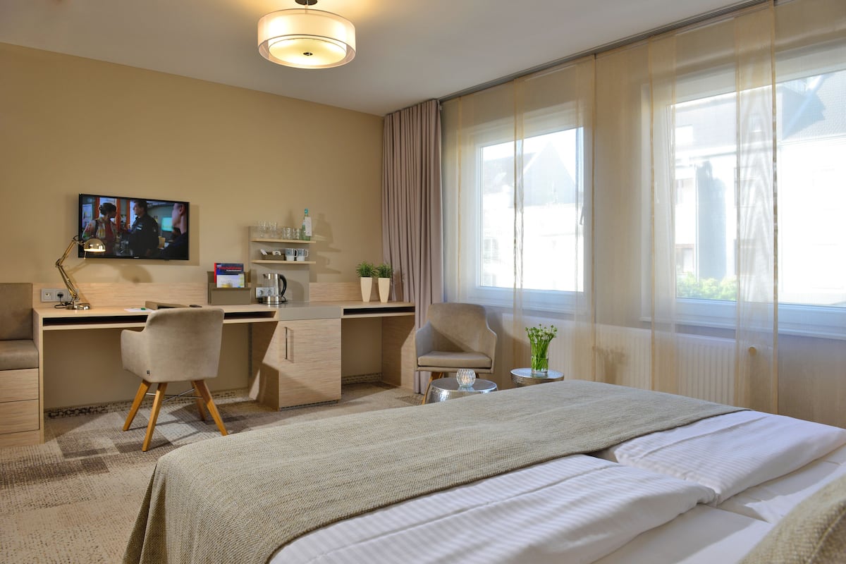 Quarto de hotel com cama, escrivaninha, cadeira, televisão e grandes janelas.