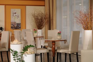 Uma área de refeições com cadeiras brancas, uma mesa de madeira e vasos com galhos secos e flores.