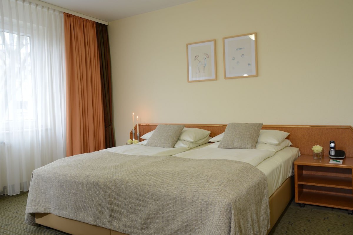 Quarto de hotel com uma cama grande, duas almofadas, uma mesa de cabeceira com luminária e duas quadros decorativos na parede.