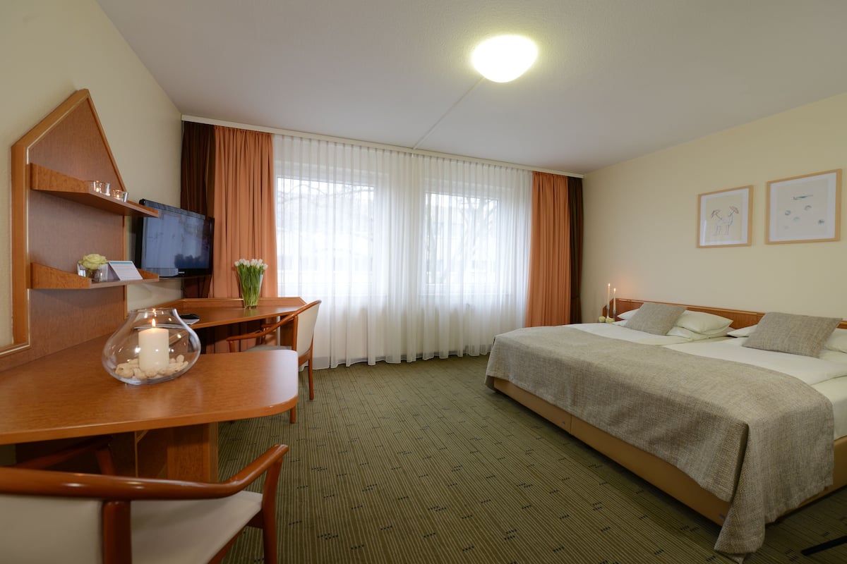 Quarto de hotel com cama, escrivaninha com televisão, vaso com flores e janela com cortinas.