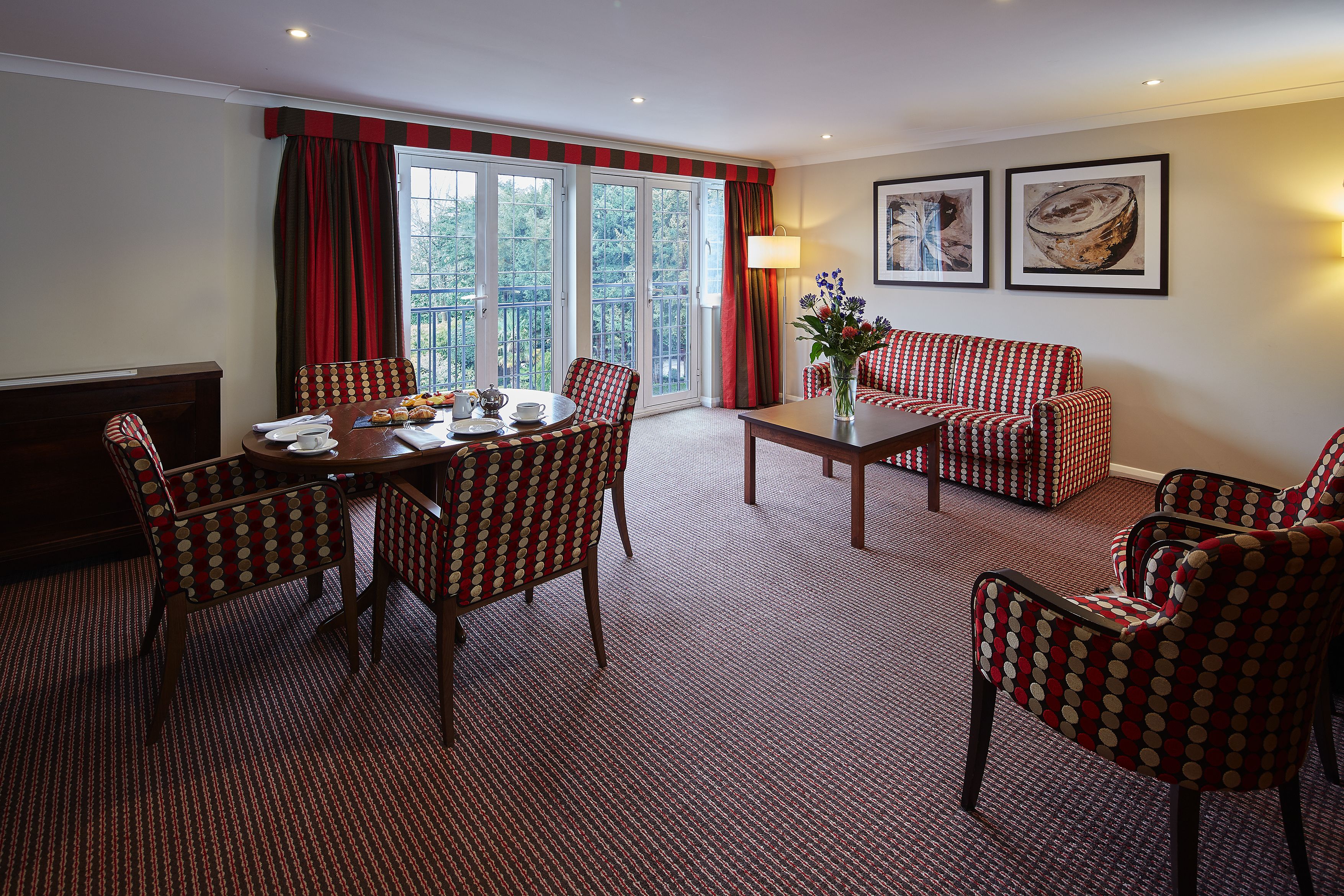 Quarto de hotel com área de jantar, um sofá, uma mesa com um vaso de flores e uma janela grande com cortinas.