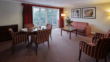 Quarto de hotel com área de jantar, um sofá, uma mesa com um vaso de flores e uma janela grande com cortinas.