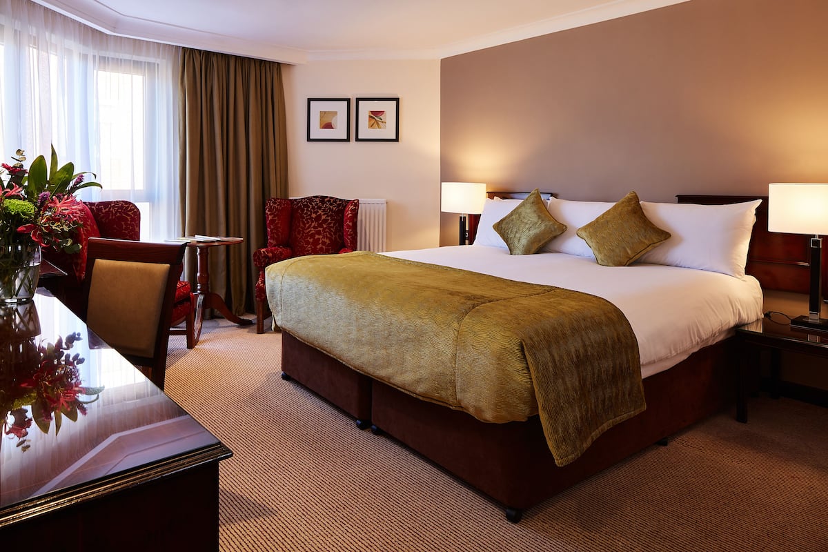 Quarto de hotel com uma cama grande, duas luminárias de cabeceira, uma escrivaninha com cadeira, uma poltrona vermelha e um vaso de flores.