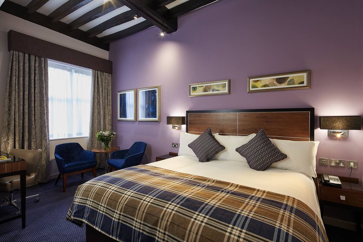 Quarto de hotel com uma cama, duas cadeiras, uma mesinha com um vaso e um criado-mudo com um telefone.