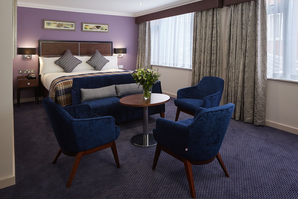 Quarto de hotel com cama, duas poltronas azuis, uma mesa redonda pequena com um vaso de flores e vista para o exterior através de cortinas finas.