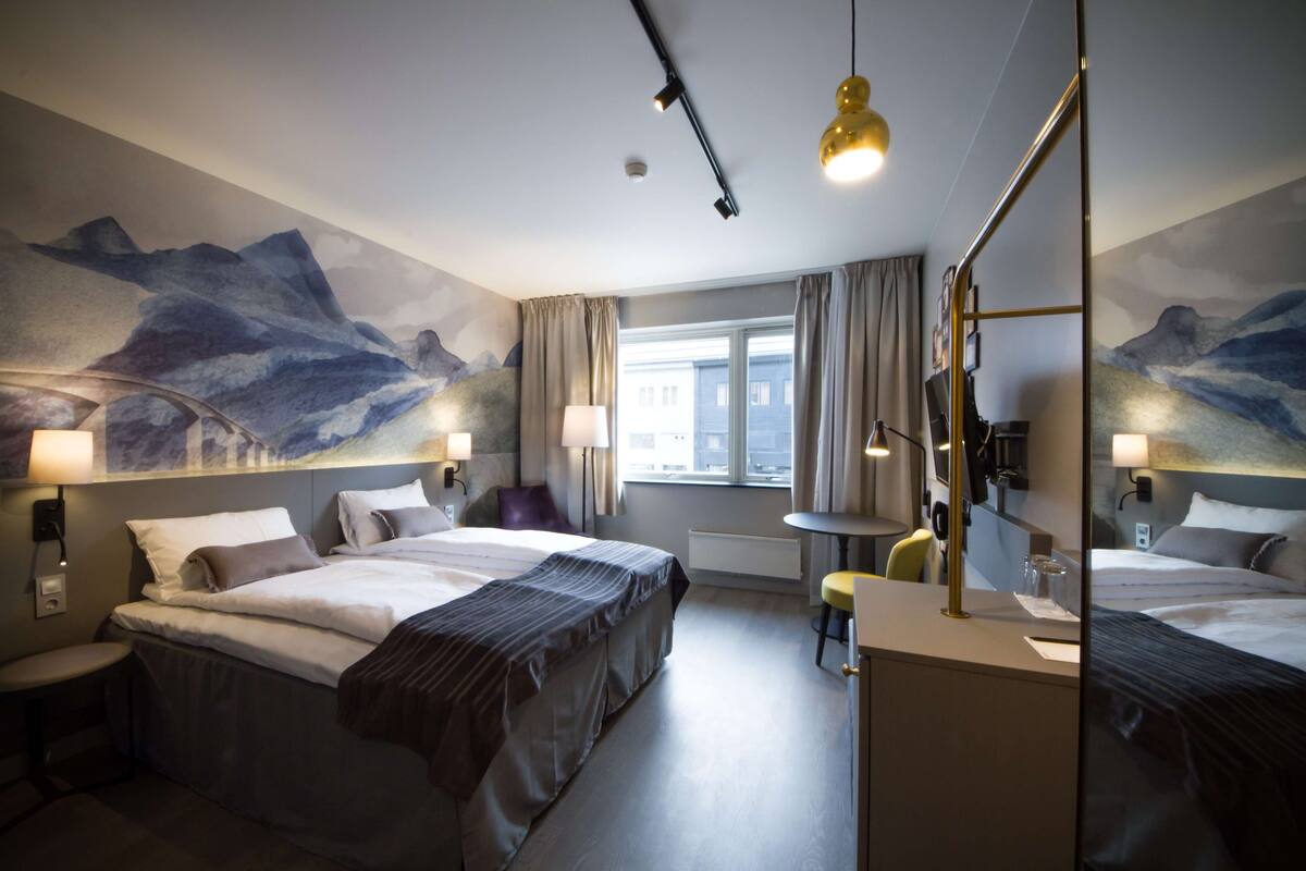 Quarto de hotel com duas camas, uma escrivaninha, uma cadeira, um espelho e uma janela com cortinas.