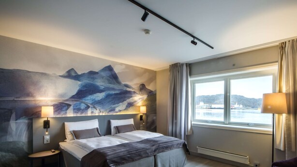 Quarto de hotel com uma cama grande, duas mesas de cabeceira, uma janela com vista para o mar e um quadro na parede.
