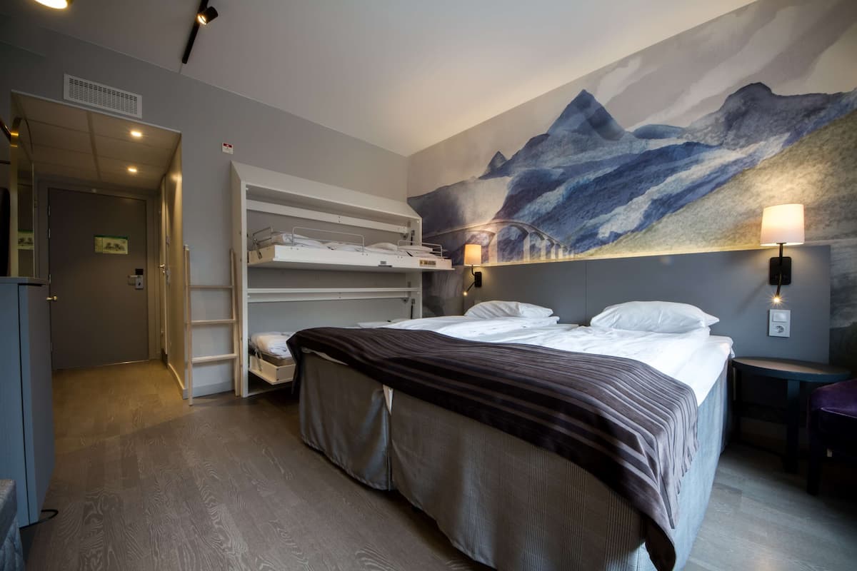 Quarto de hotel com beliche, pintura de montanhas na parede, escrivaninha com abajur e armário.