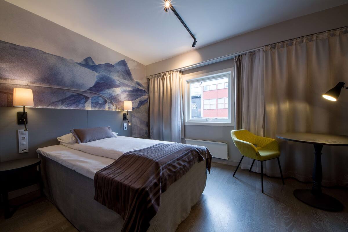 Quarto de hotel com uma cama, uma cadeira, uma mesa, um abajur, um quadro e uma janela com cortinas.
