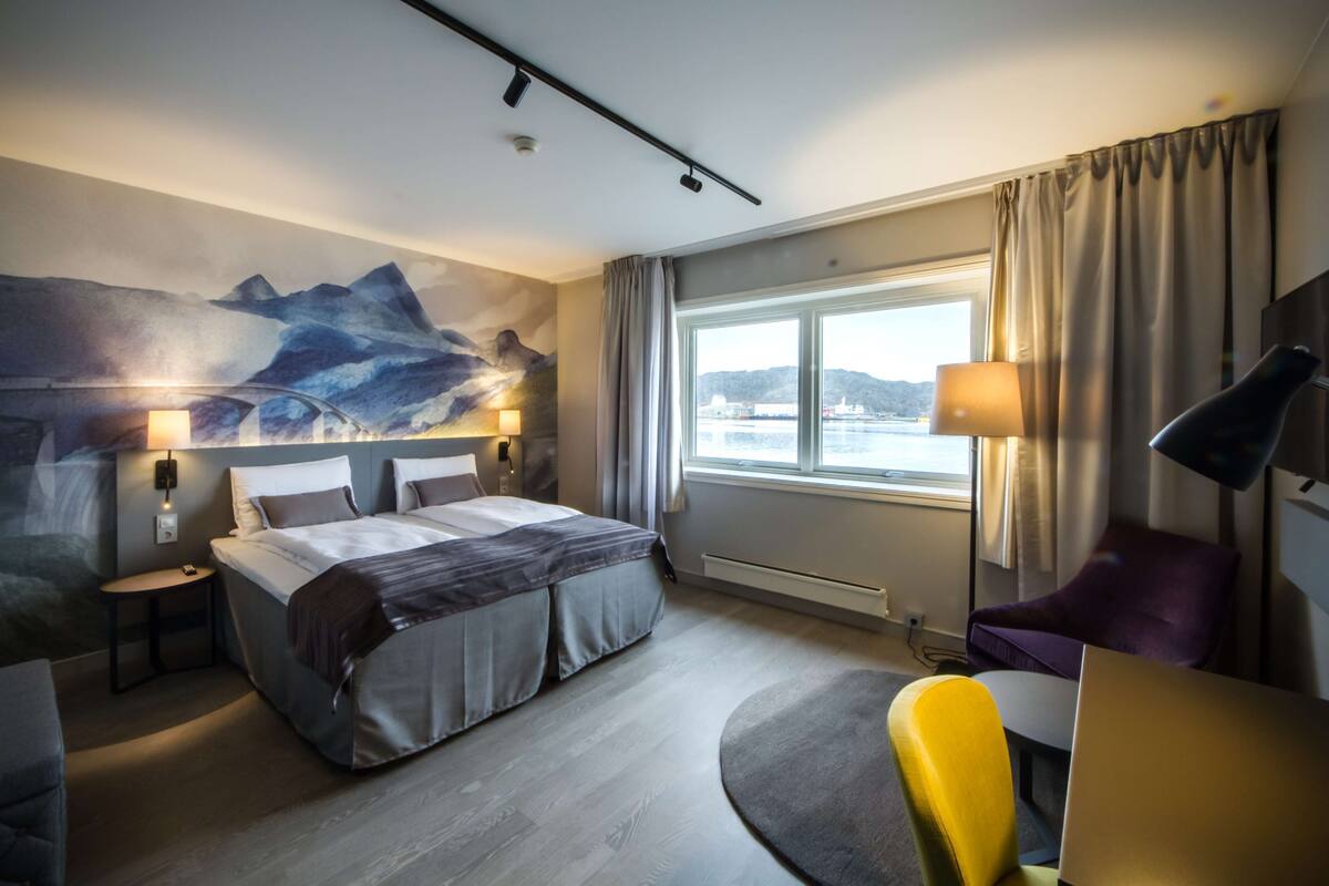 Quarto de hotel com cama, escrivaninha, cadeira, janela com vista para um corpo de água e um quadro na parede.