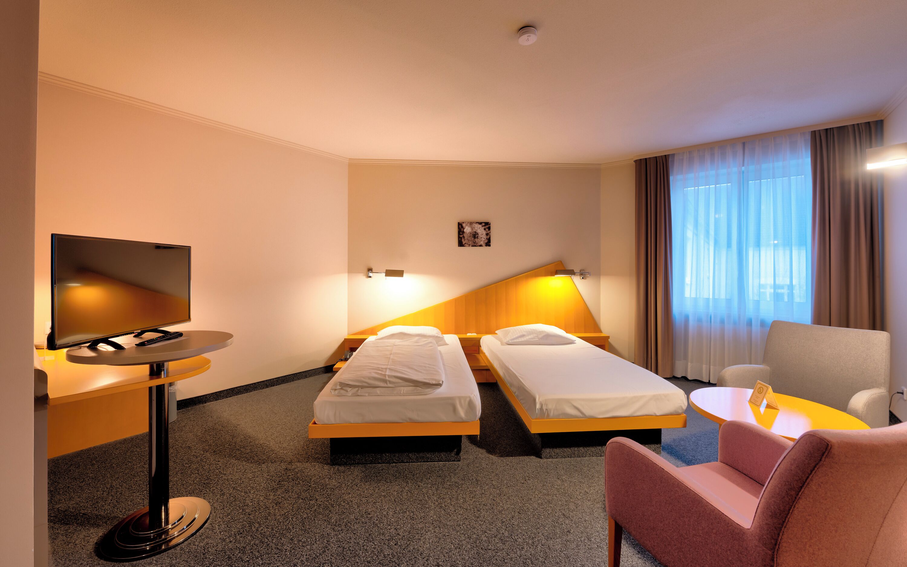Quarto de hotel com duas camas de solteiro, uma televisão, uma escrivaninha com cadeira e uma mesinha.