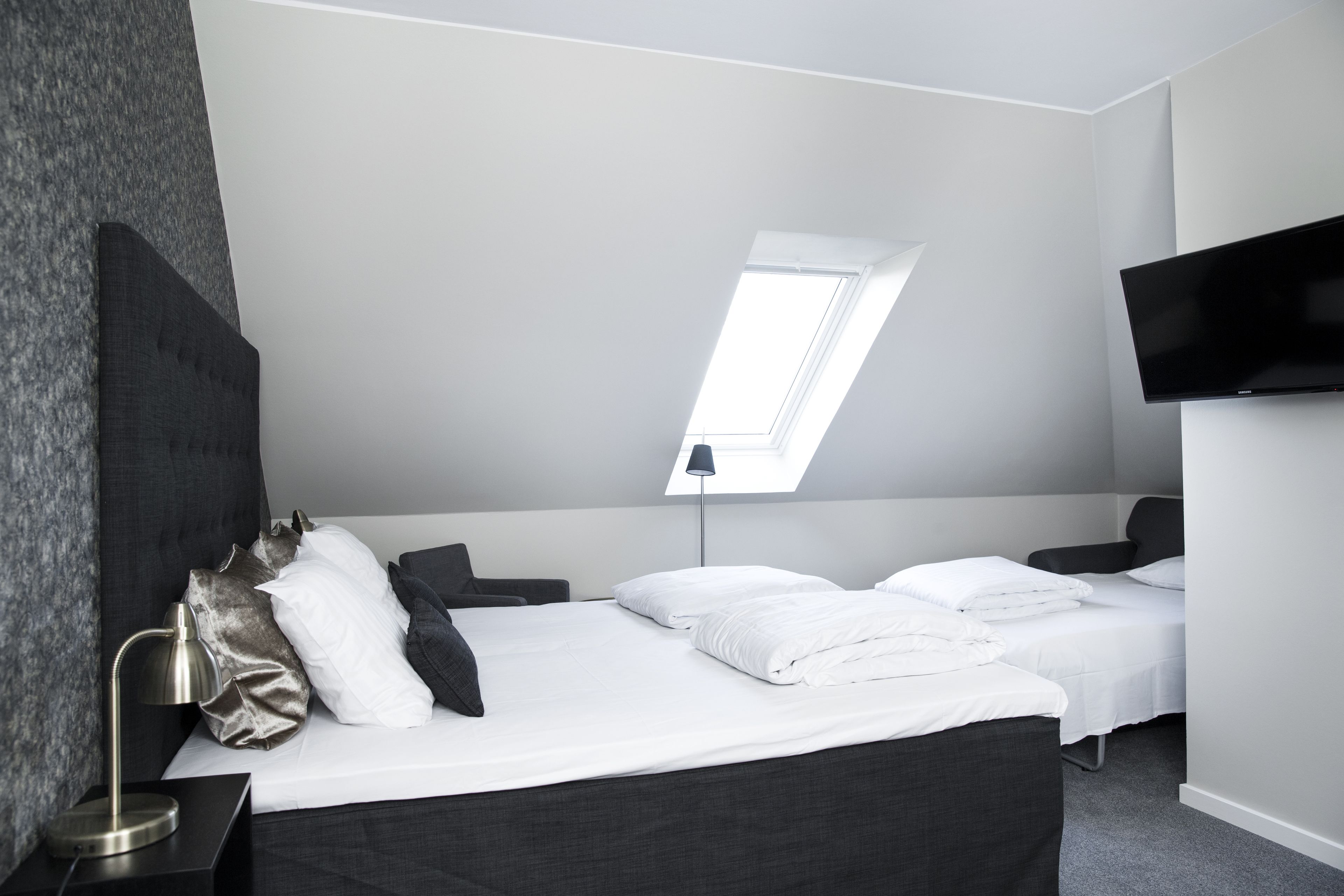 Quarto de hotel moderno com uma cama grande, uma cama menor, uma TV de tela plana, uma escrivaninha com abajur e um claraboia.