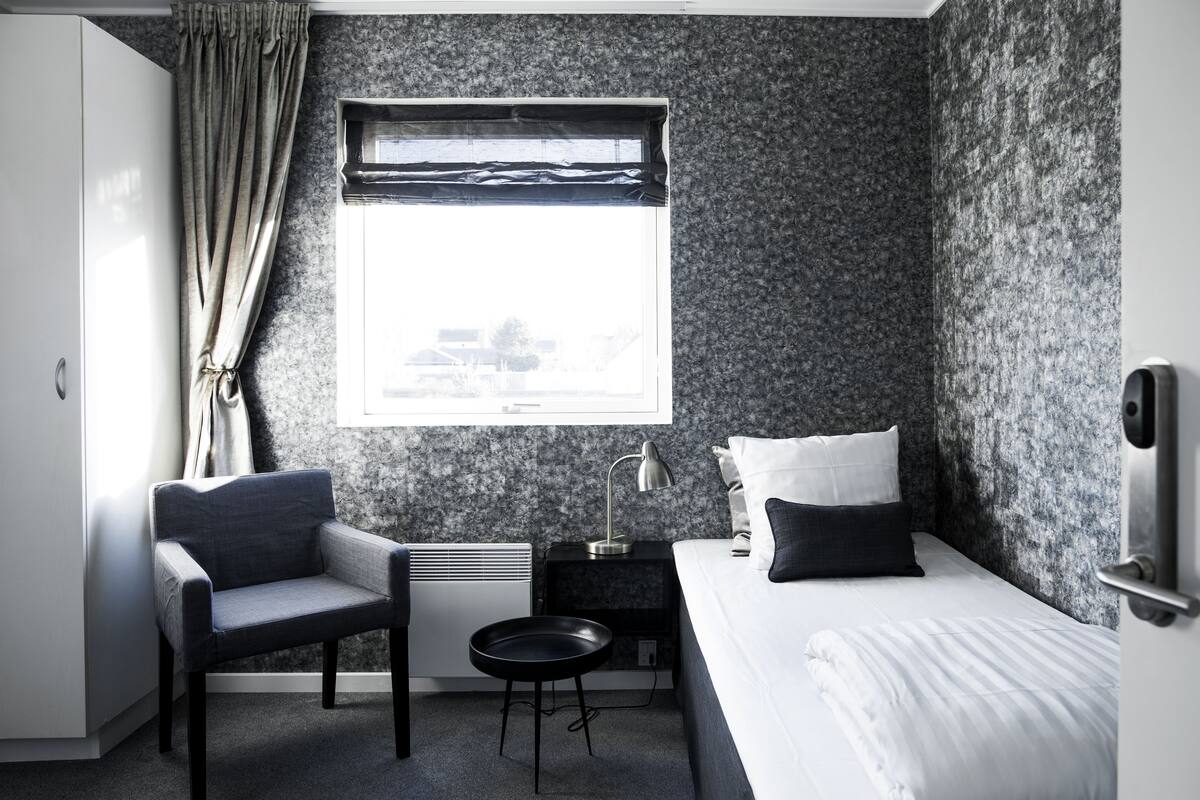 Quarto de hotel moderno com cama, uma cadeira, uma mesinha e janela com cortinas.