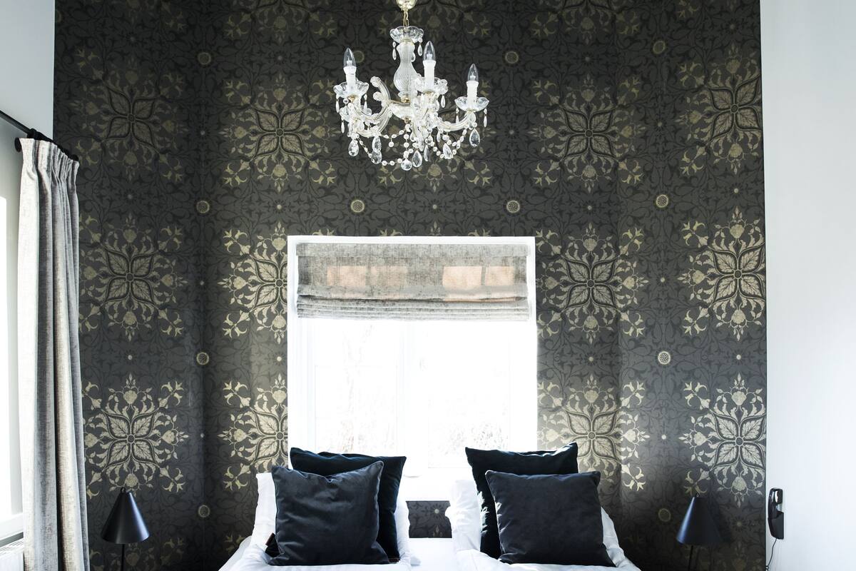 Quarto com cama grande, papel de parede estampado escuro, um lustre e uma janela.
