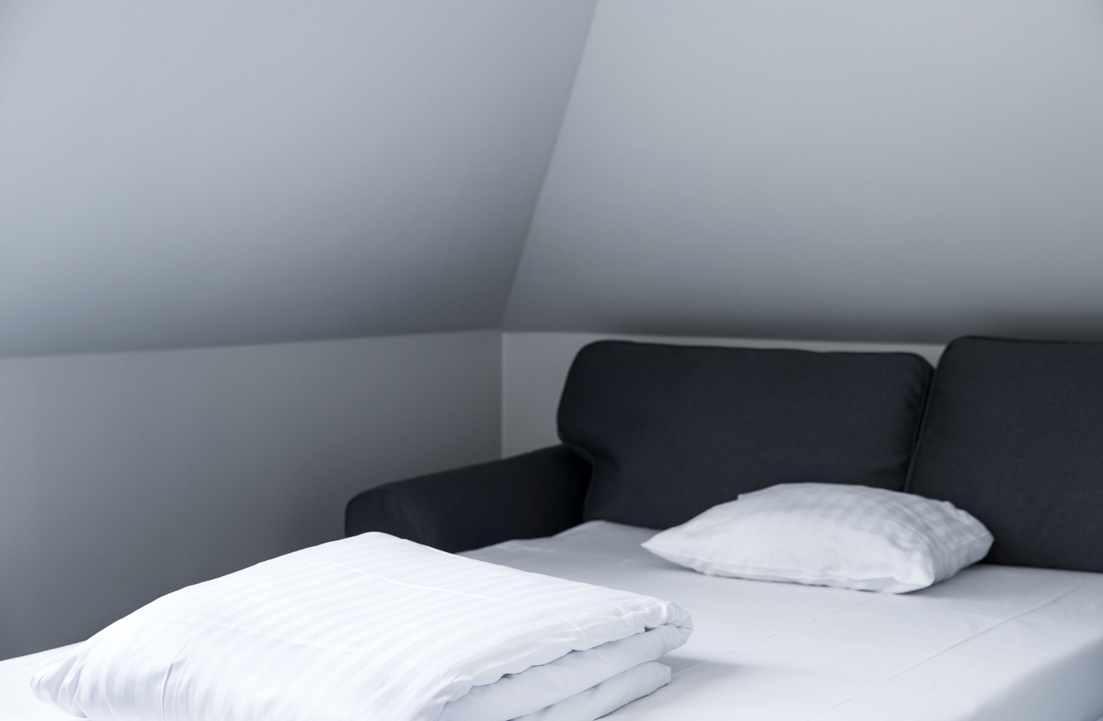 Quarto moderno e minimalista, com um sofá escuro e uma cama bem arrumada com roupa de cama branca.