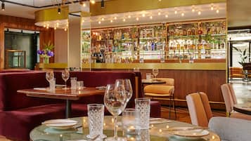 Uma área de bar com diversas garrafas de bebidas, um espaço de estar com mesas e cadeiras, e um lustre decorativo.