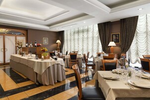 Uma área de refeições de hotel com mesas preparadas para o serviço, um bar com garrafas e vista para o quarto através de cortinas.