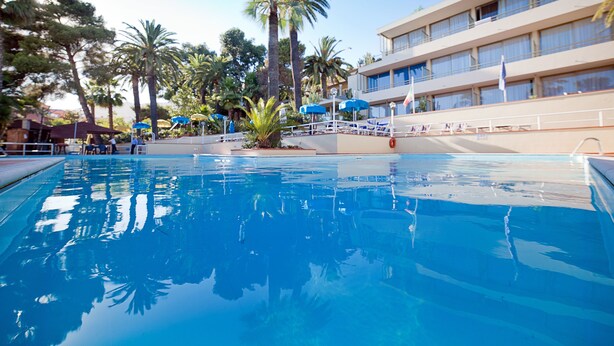 Piscina cercada por um prédio de hotel com várias varandas, com palmeiras e guarda-sóis.