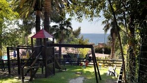 Um parquinho com baloiço e um pequeno abrigo, rodeado por árvores e vista para o mar.