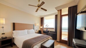 Quarto de hotel com cama, mesa de cabeceira, abajur, escrivaninha, cadeira e televisão.