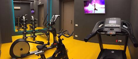 Academia com equipamentos de cardio, um espelho e uma TV exibindo um artista.