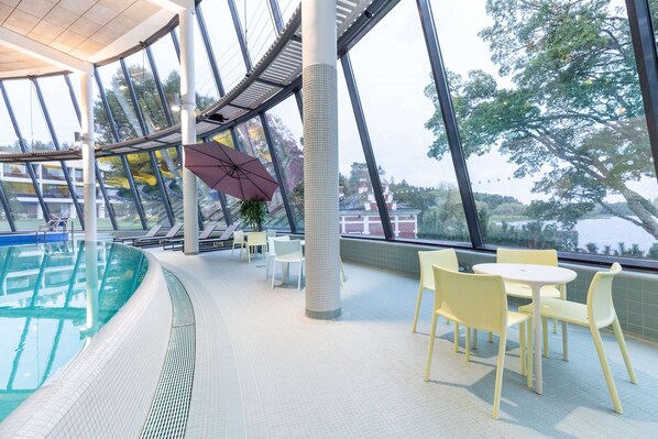 Uma piscina interior com paredes de vidro transparente, cadeiras amarelas e uma mesa com guarda-sol vermelho.