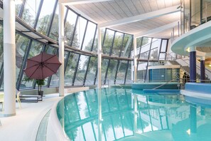Uma grande piscina interior com paredes de vidro transparente, um guarda-sol e uma escadaria que leva a um andar superior.