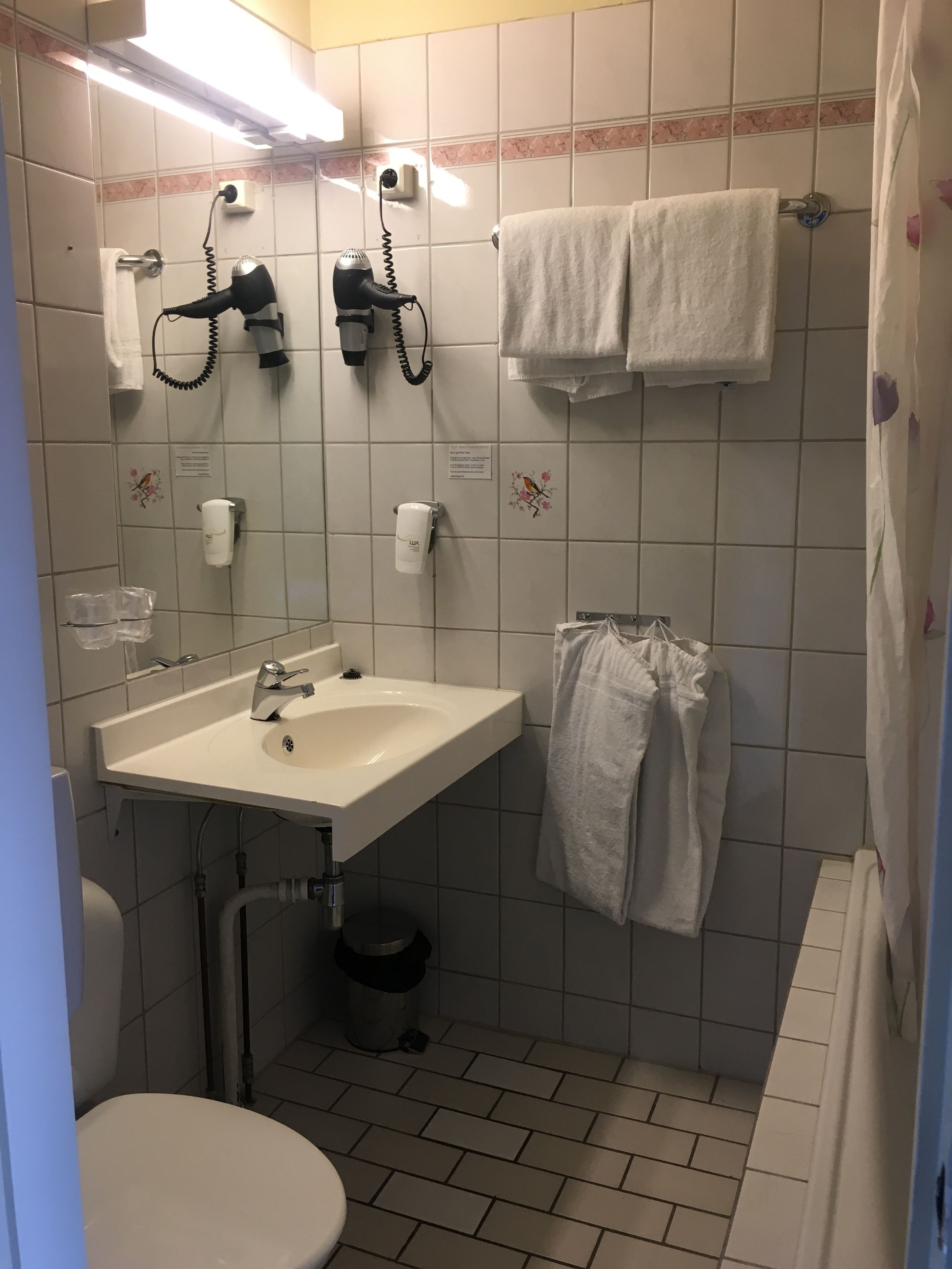 Uma casa de banho pequena com uma pia, dois secadores de cabelo, sanita e cortina de chuveiro.