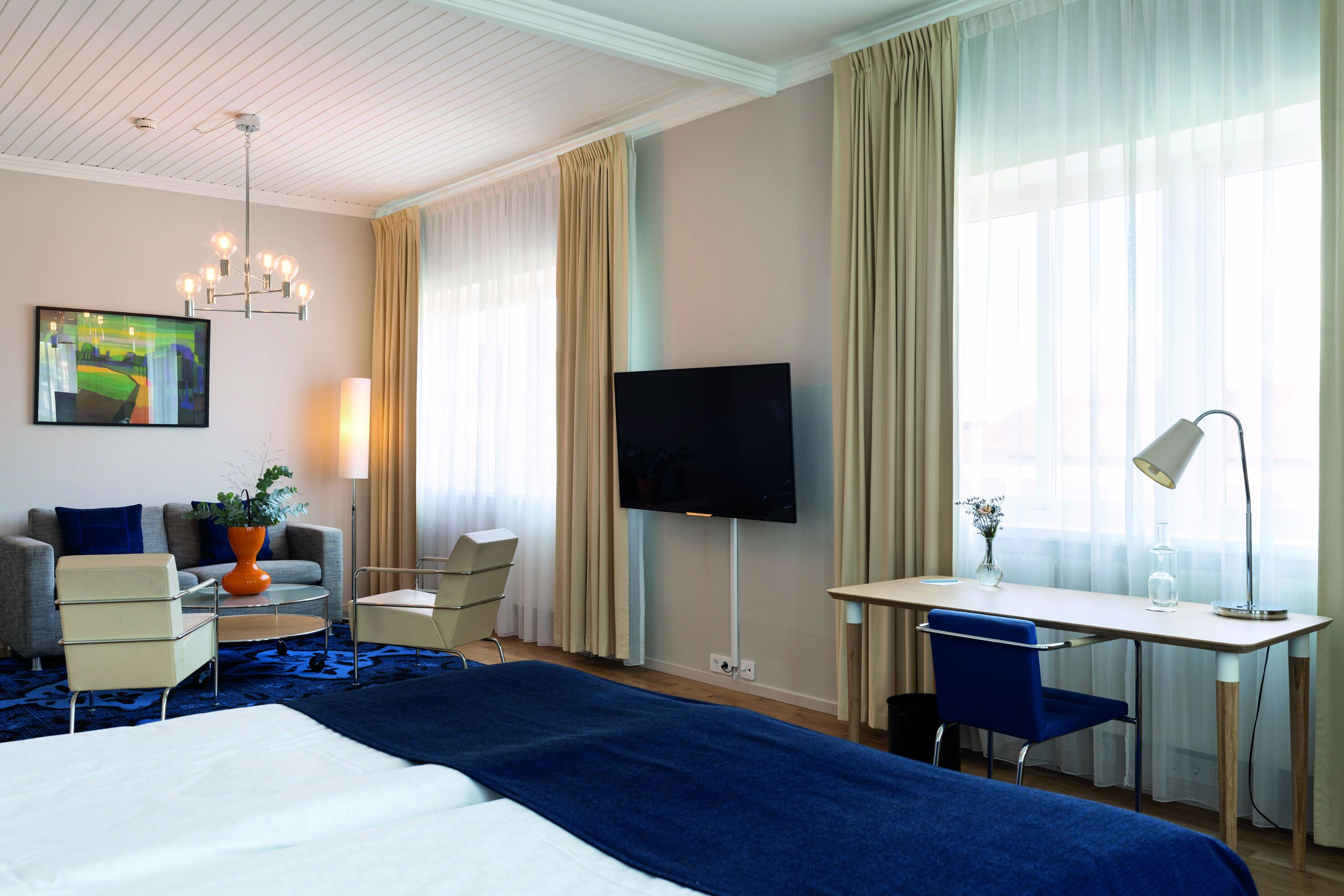 Quarto de hotel com cama, escrivaninha, televisão, sofá e poltrona.