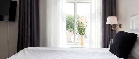 Quarto de hotel com uma cama, televisão fixada na parede, mesa de cabeceira com luminária e uma janela com cortinas.
