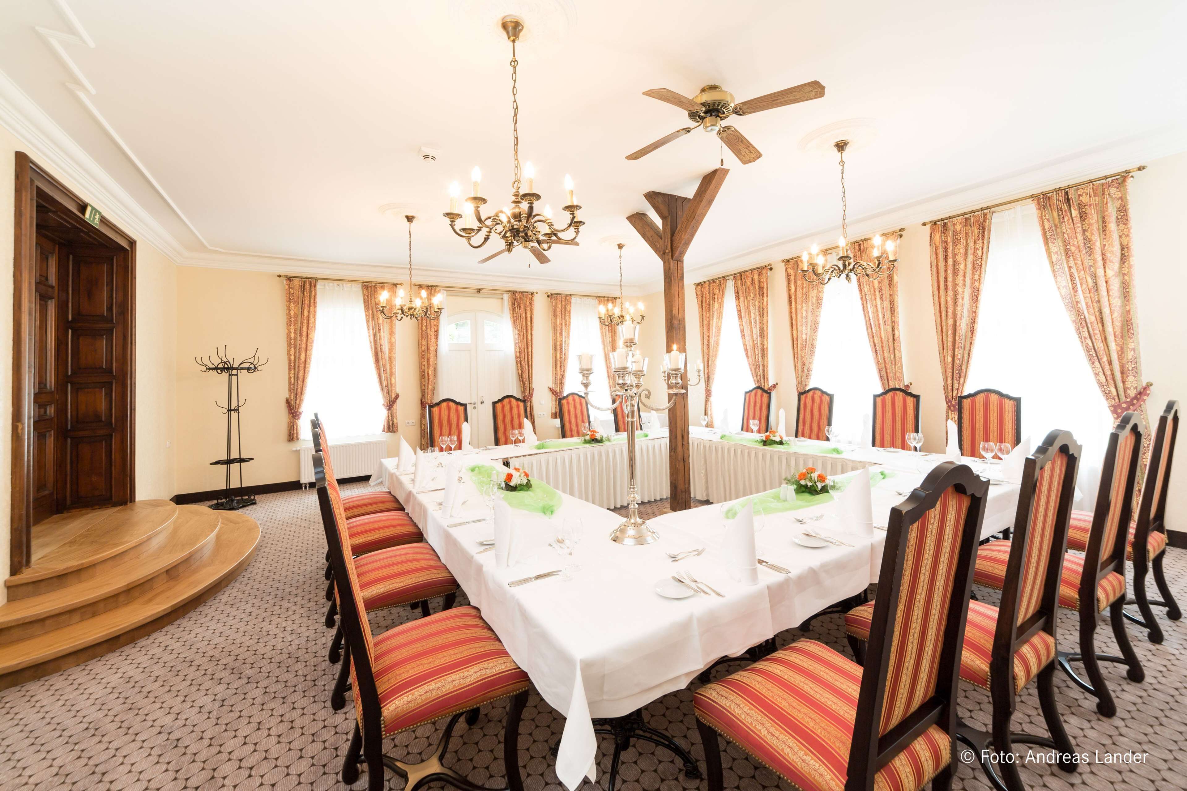 Uma sala de jantar formal com uma mesa comprida, cadeiras, lustres e um ventilador de teto.