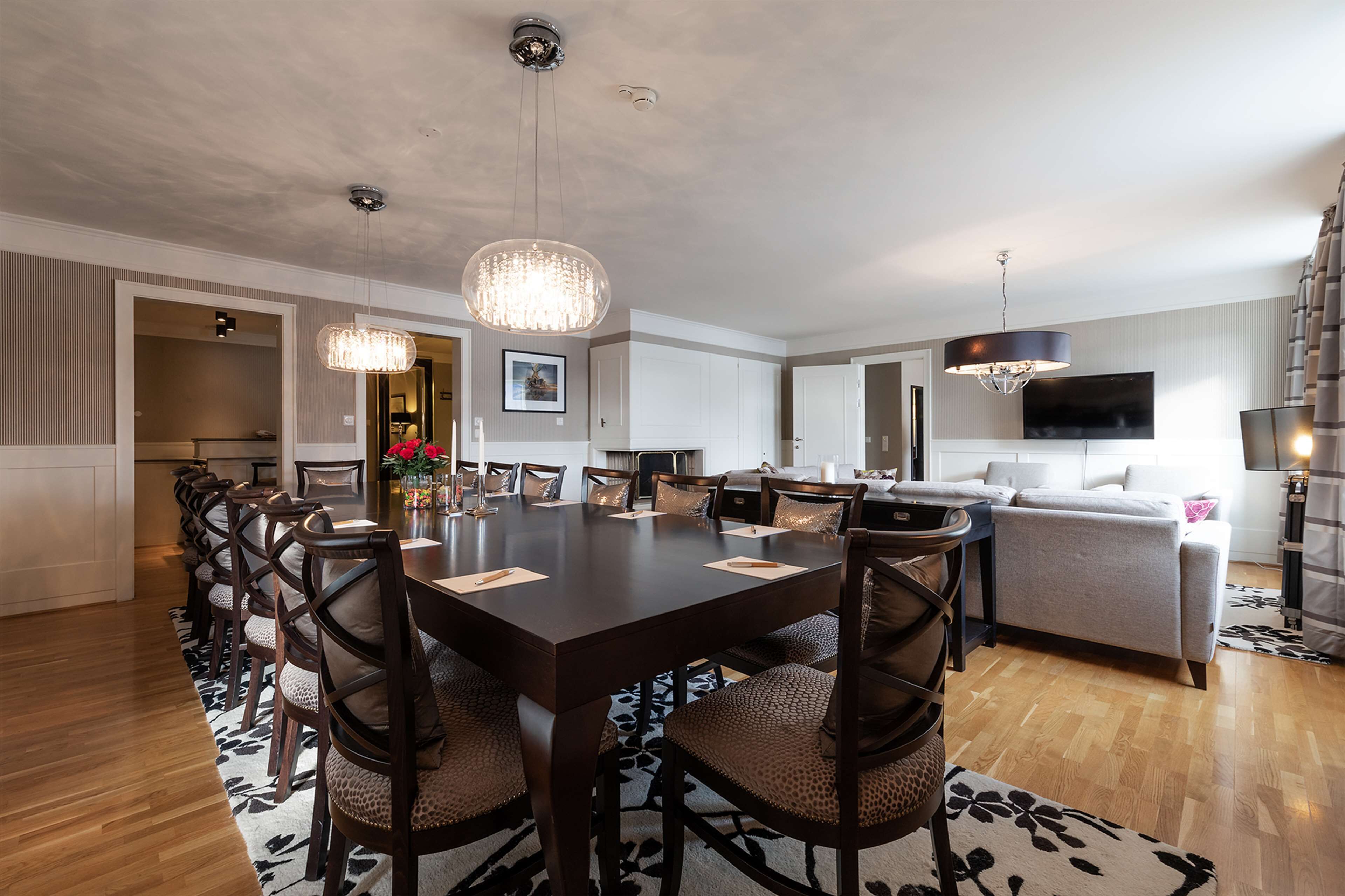 Uma sala de jantar moderna com uma mesa comprida e cadeiras, um candelabro e um sofá ao fundo.