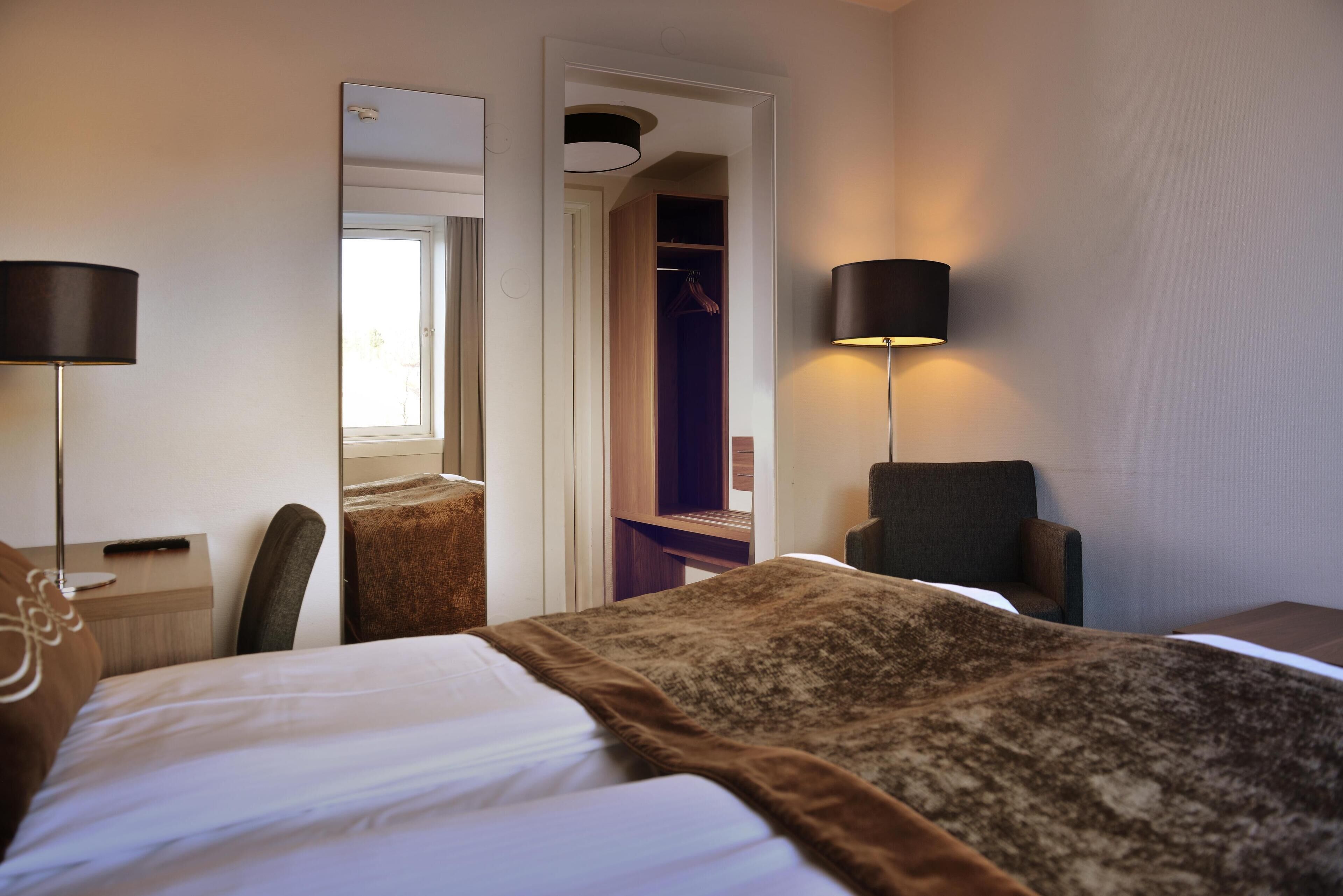 Quarto de hotel com cama, luminárias de cabeceira, uma cadeira, escrivaninha com controle remoto e guarda-roupa.