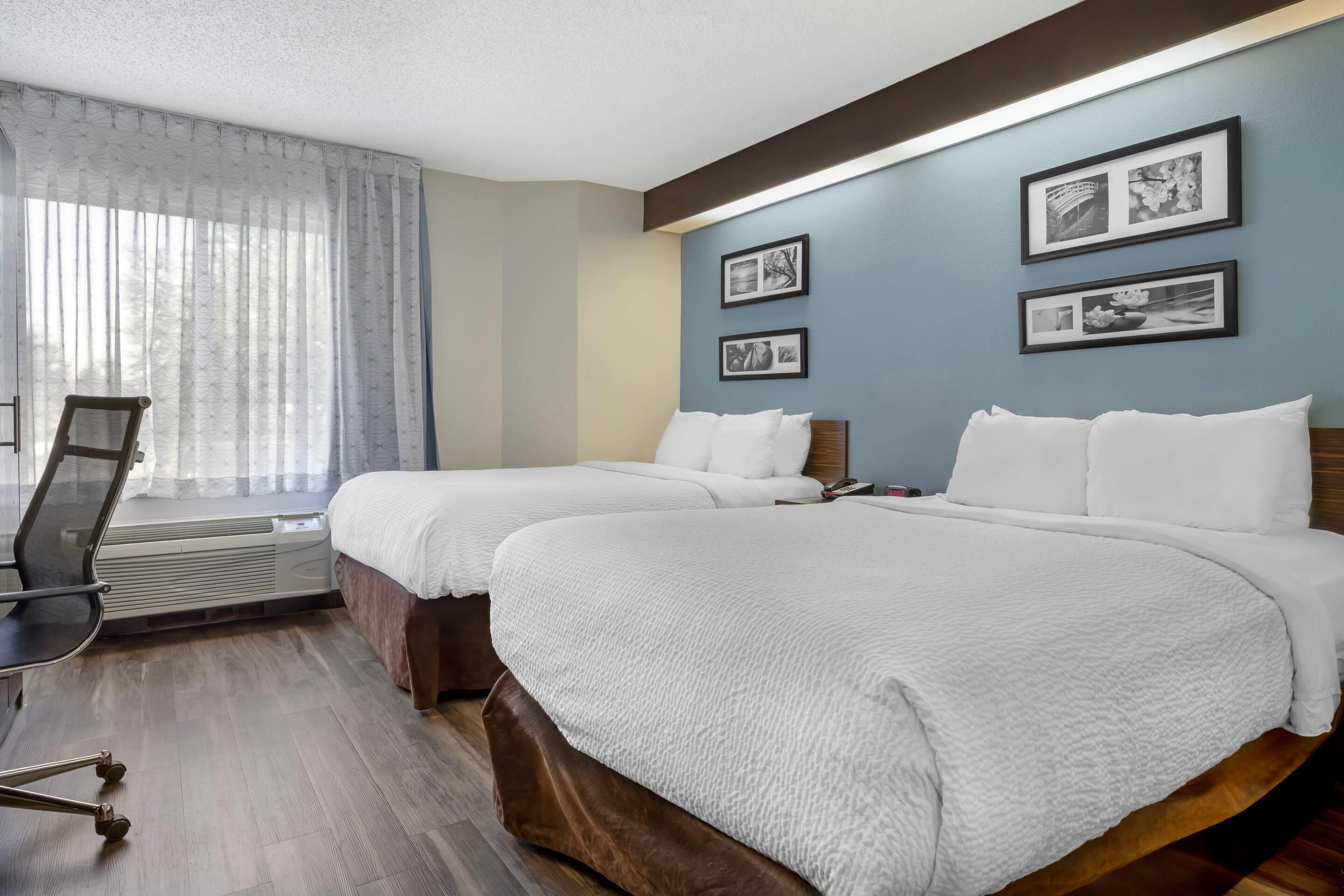 Quarto de hotel com duas camas, uma escrivaninha, uma cadeira e uma janela grande com cortinas.