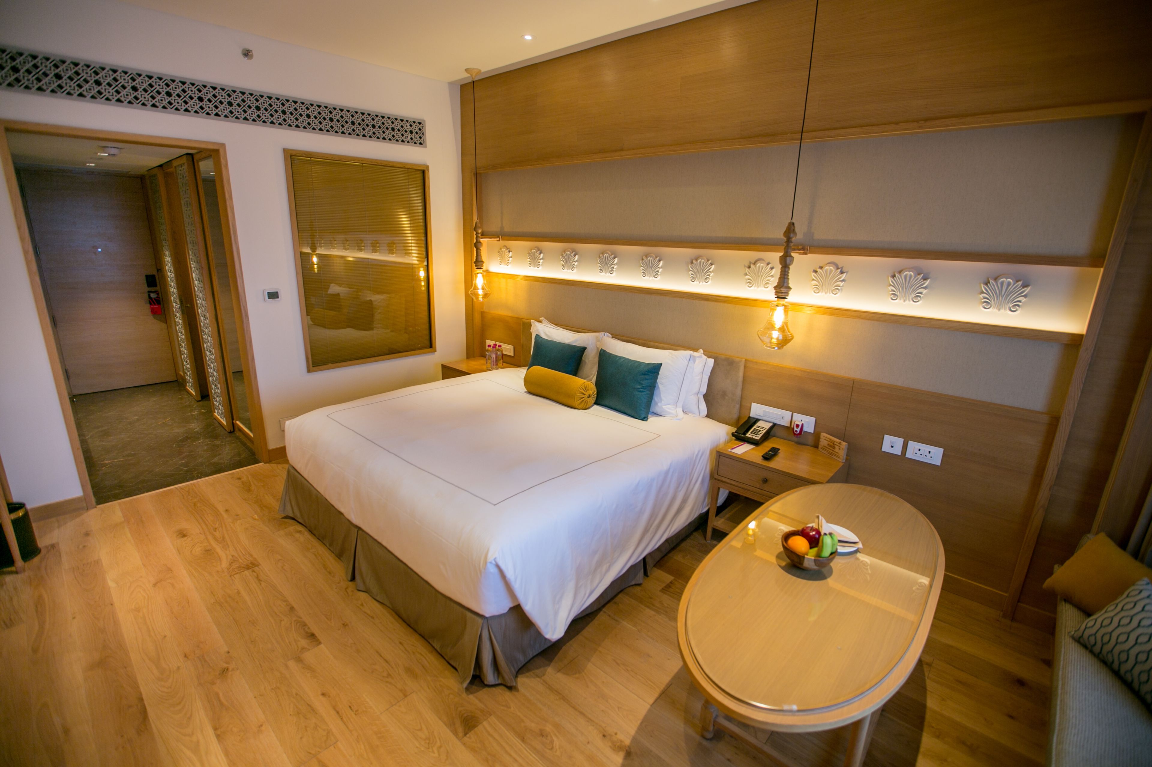 Quarto de hotel com uma cama grande, mesa de cabeceira e uma pequena mesa com frutas.