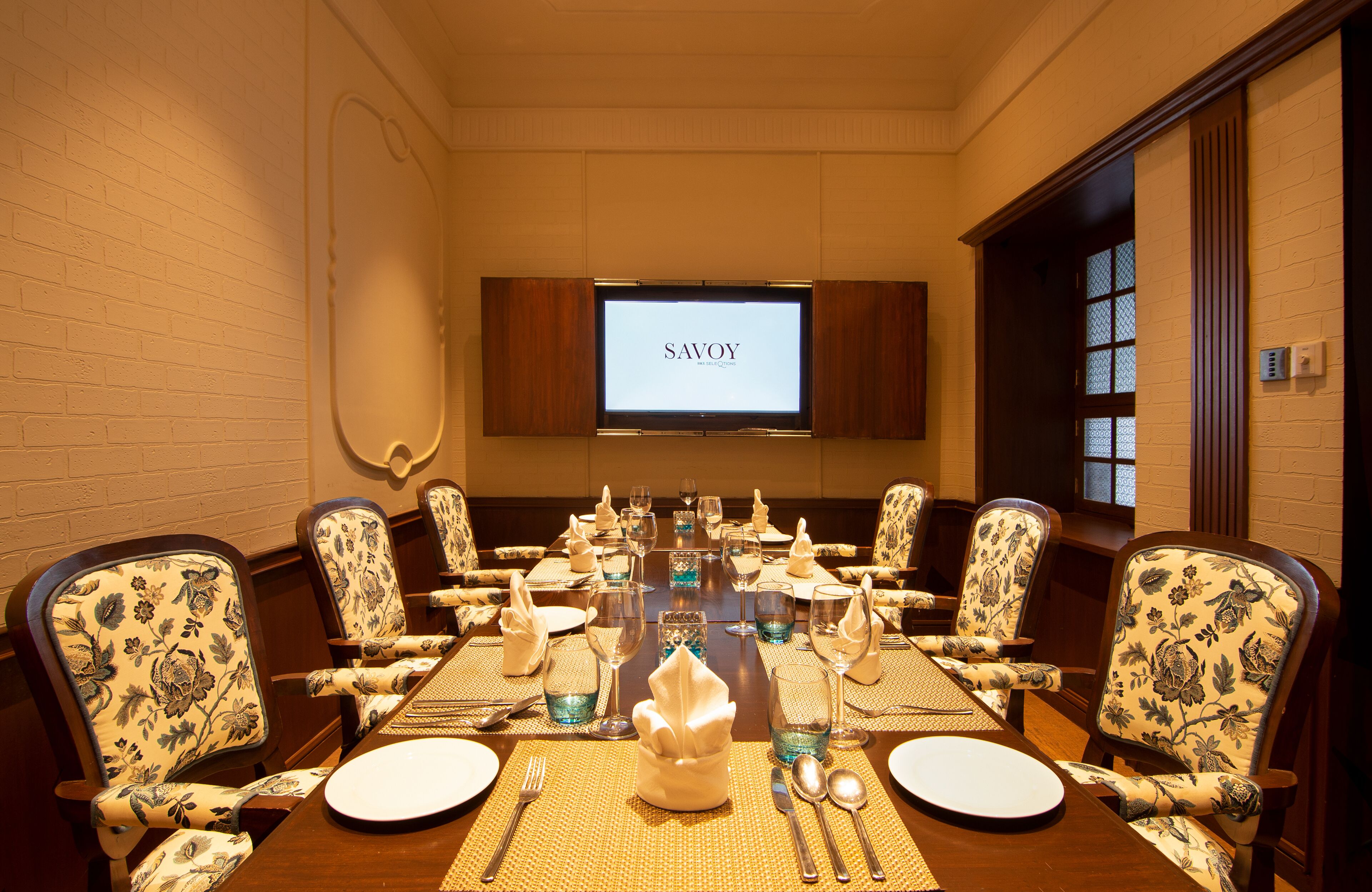 Uma sala de conferências com uma mesa comprida, cadeiras com padrão floral e uma tela exibindo "SAVOY" na parede.