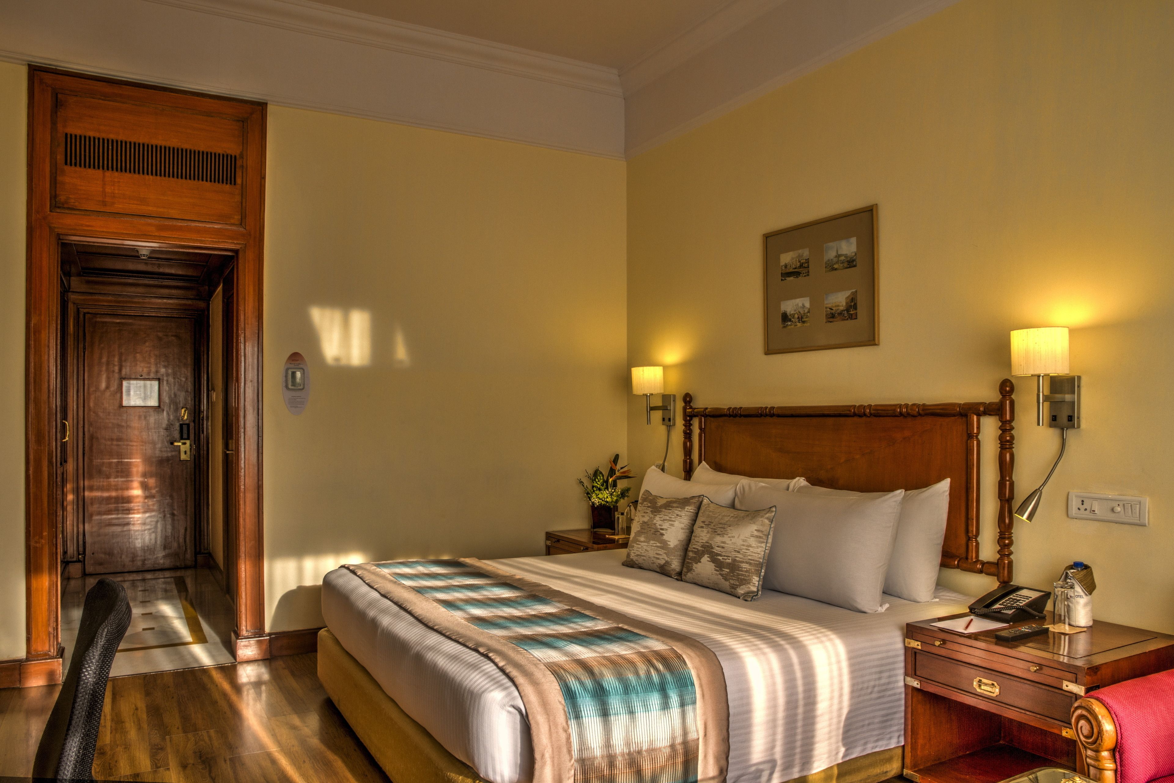 Quarto de hotel com uma cama grande, cabeceira de madeira, duas mesas de cabeceira com luminárias, um criado-mudo e uma cadeira vermelha.