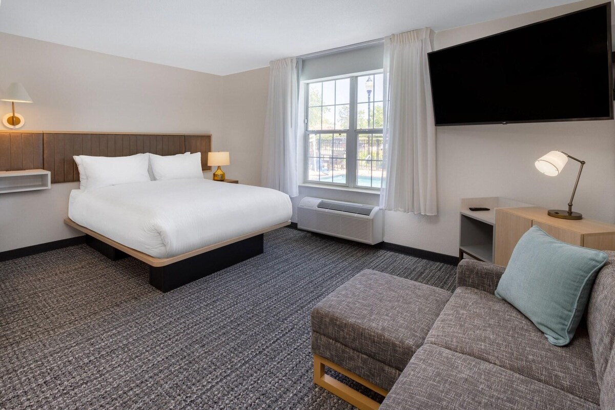 Quarto de hotel com uma cama grande, um sofá, uma televisão e uma janela com cortinas.