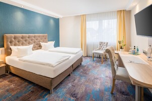 Quarto de hotel com uma cama grande, escrivaninha, cadeira e televisão.