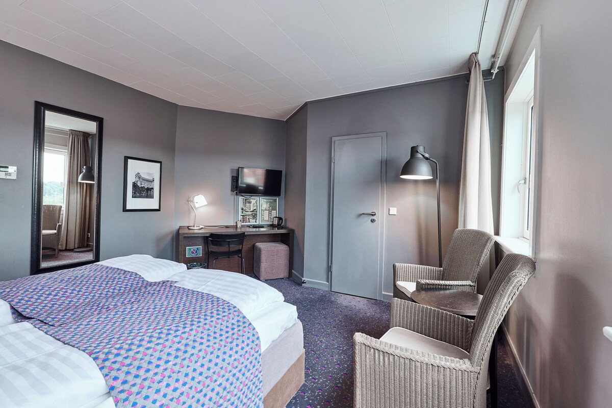 Quarto de hotel com uma cama, escrivaninha, cadeira, televisão e porta.