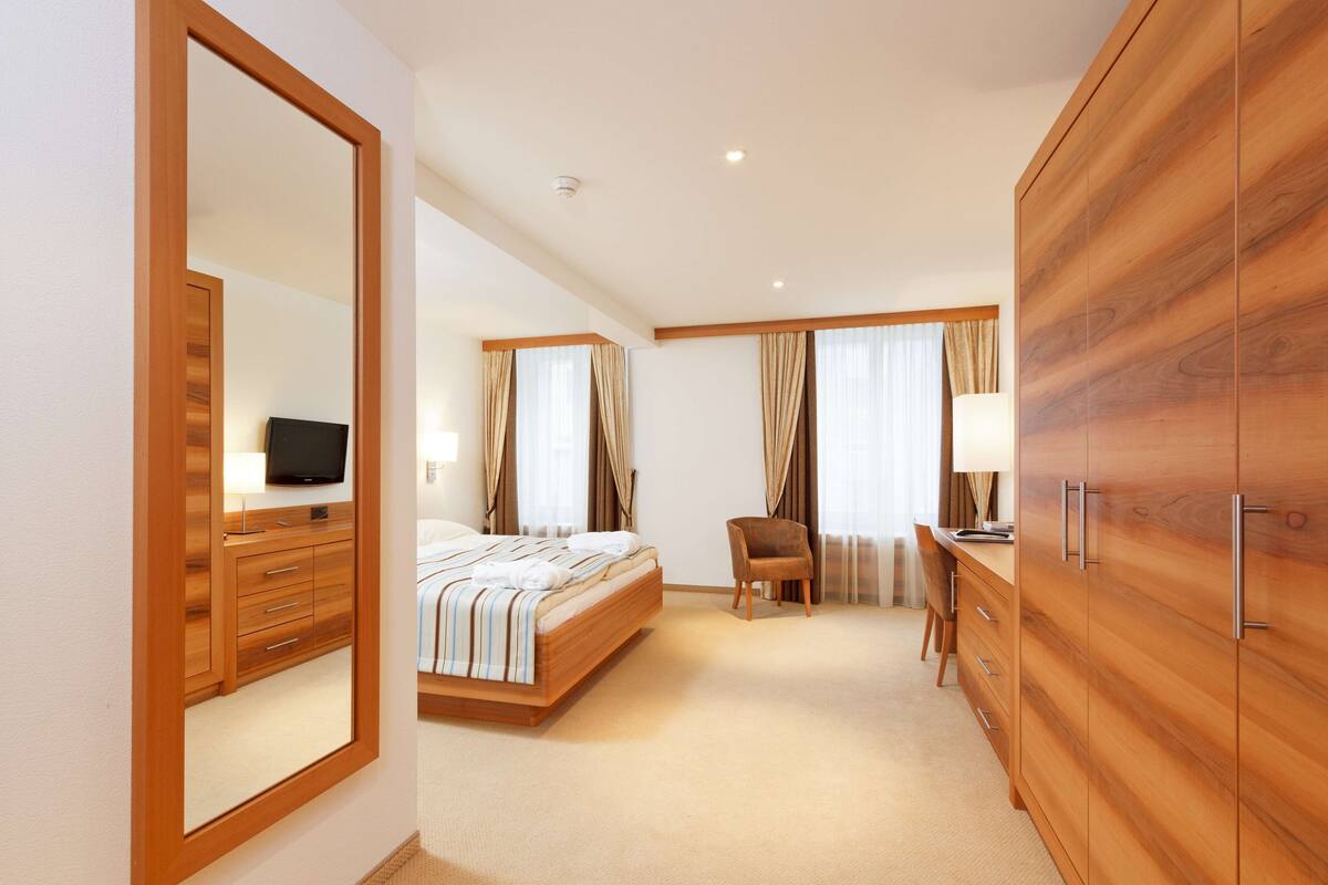 Quarto de hotel com guarda-roupa de madeira, cama com manta listrada, escrivaninha com luminária e uma cadeira.