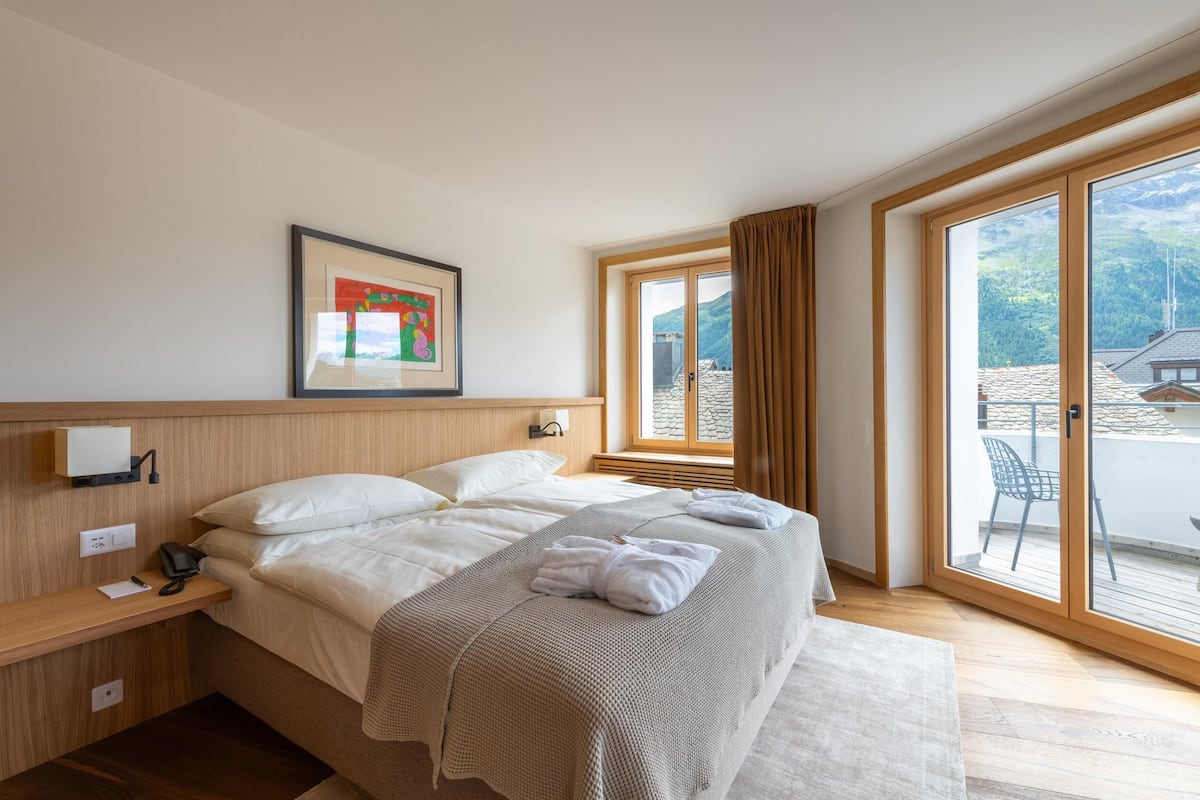 Quarto de hotel com uma cama, mesa de cabeceira com telefone e luminária, quadro pendurado na parede e uma grande janela com vista para o exterior.