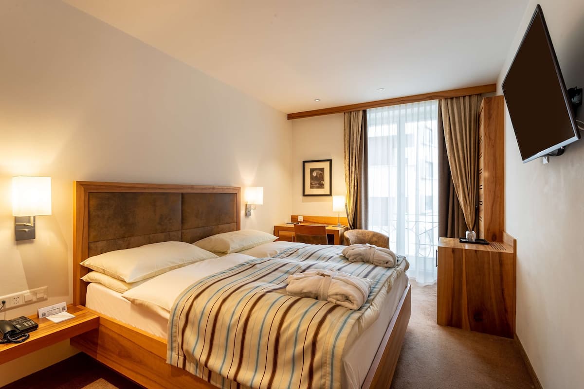 Quarto de hotel com cama, mesa de cabeceira, luminária, escrivaninha, cadeira e televisão fixada na parede.