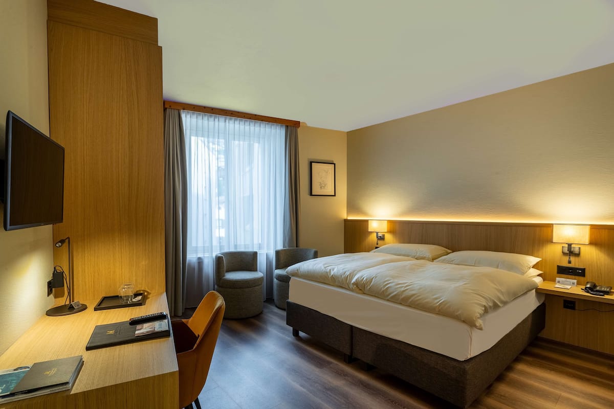 Quarto de hotel com cama, mesas de cabeceira, escrivaninha, cadeira, televisão e janela com cortinas.