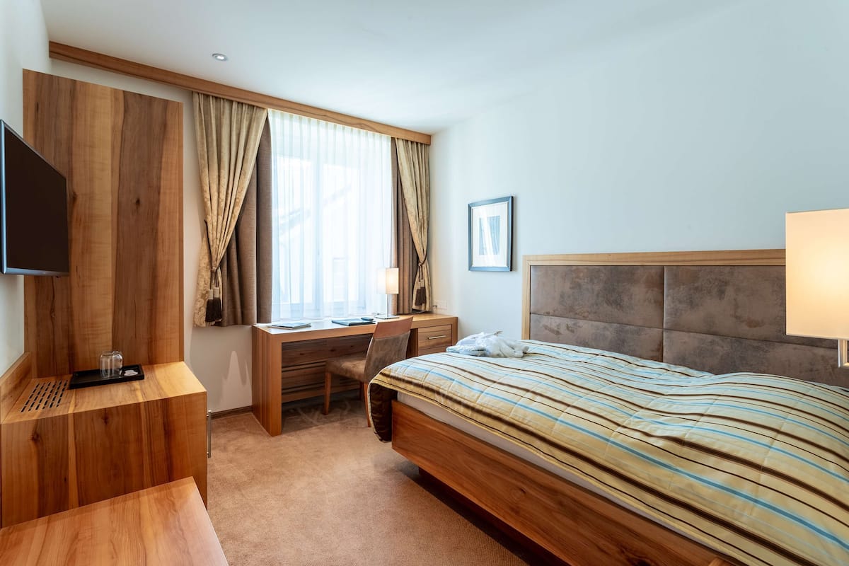 Quarto de hotel com escrivaninha de madeira, cama com roupa de cama listrada, televisão e janela com cortinas.